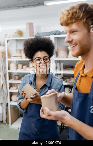 cheerful interracial couple molding clay cups during date in pottery ...