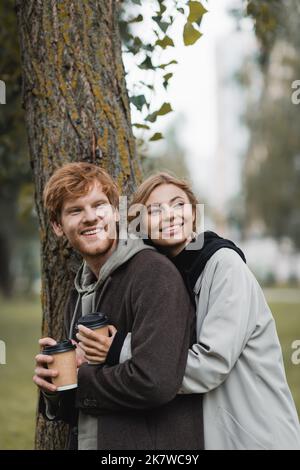 happy blonde woman holding paper cup and leaning on shoulder of smiling ...