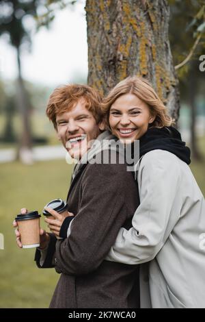 cheerful blonde woman holding paper cup and hugging with boyfriend near ...