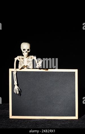 Funny human skeleton holding an empty black blank on a black background ...