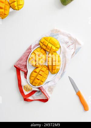 A flat lay of cut mangoes, a knife and pink flowers on a white ...