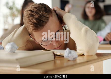 Youthful schoolboy bending over desk and covering his head by hands ...