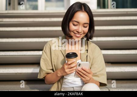 Happy urban girl drinks her takeaway coffee and scrolls feed on ...