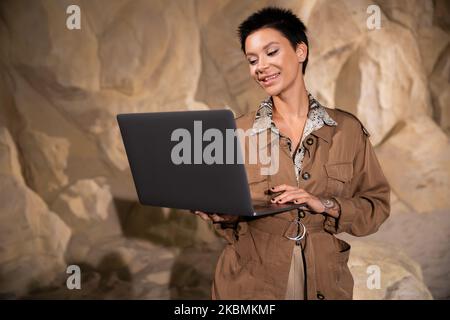 happy archaeologist with tattoo and short hair using laptop in cave ...