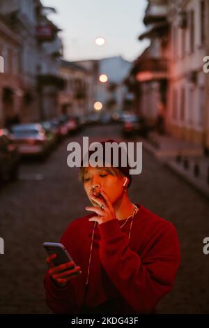 Asian boy in earphones using mobile phone while smoking at city street ...