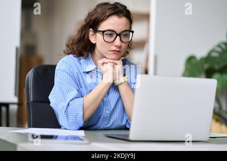 Serious young woman watching webinar on laptop and writing in notebook ...