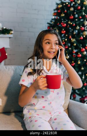 Kid in pajama talking on smartphone and touching ball on Christmas tree ...