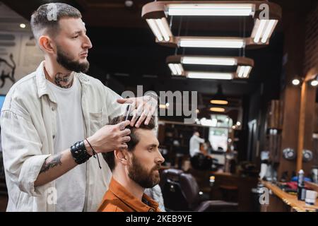 Tattooed barber combing hair of client and holding scissors in ...