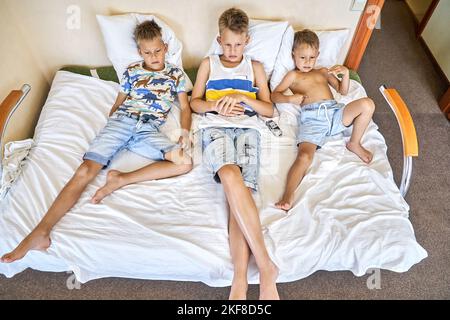 Exhausted boy siblings watch together movies on TV lying on double bed ...