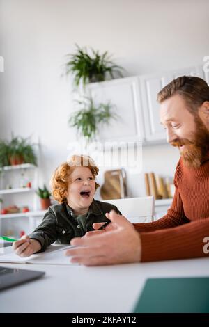 bearded man talking to redhead son doing homework near blurred laptop ...