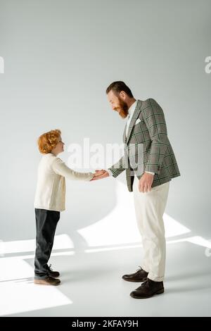 Red haired boy in jumper sitting near stylish dad on grey background ...