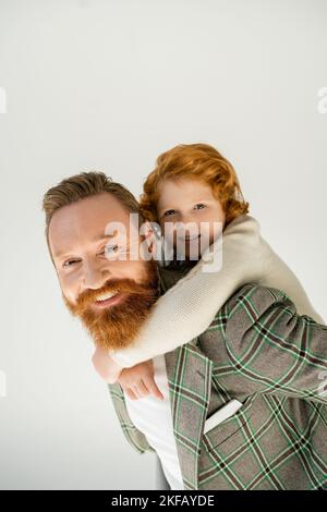 Redhead boy in knitted jumper embracing bearded dad on grey background ...