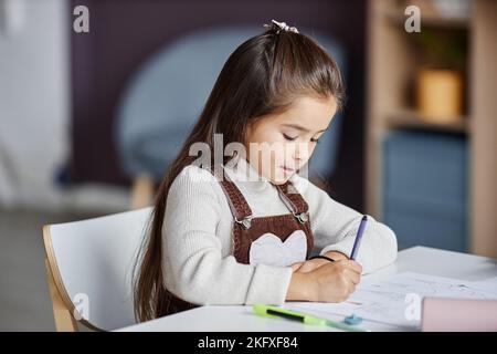 Diligent little girl with dark long hair drawing with crayons on paper ...