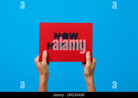 a man holds a red paper sign with the text now hiring written in black ...