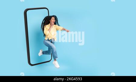 Excited woman jumping and pointing with finger at smartphone on pink ...