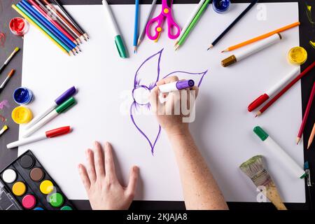 Overhead of hands drawing purple flower on paper, with art materials on ...