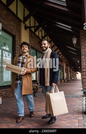 happy gay couple in scarfs looking into shopping bags near blurred ...