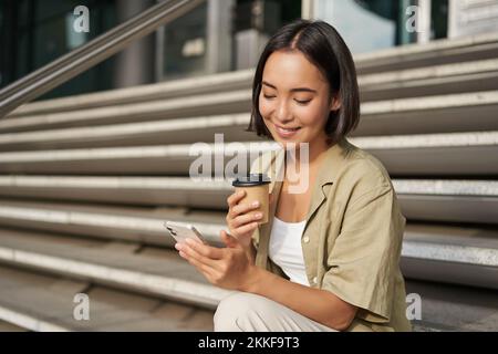 Happy urban girl drinks her takeaway coffee and scrolls feed on ...