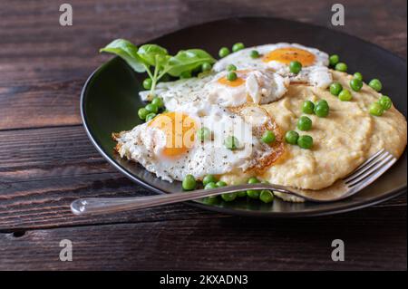 Creamy polenta with fried eggs, sunny side up and green peas on dark ...