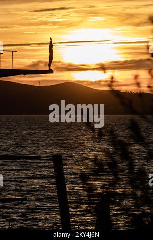 Boy jumps off the diving platform into the Adriatic Sea during sunset ...