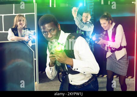 Portrait of Afro man playing laser tag with his co-workers on dark ...