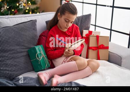 Adorable hispanic girl writing on notebook sitting on floor by ...