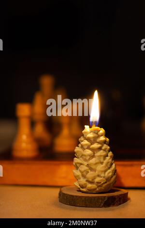 Burning candle in pine cone shape. Chess in the background. Handmade ...