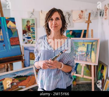 Woman painter holding paintbrush in hand and palette while smiling and ...