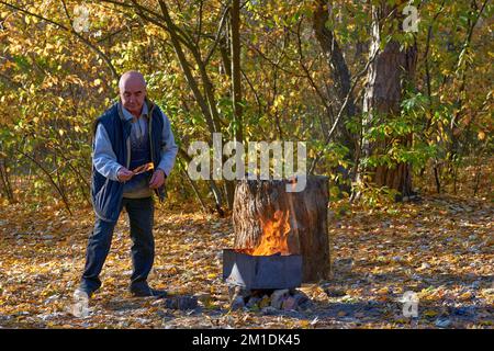 Cheerful elderly man pensioner makes a fire for grilling at a picnic in ...