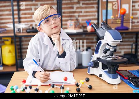 Adorable toddler student writing on notebook with doubt expression at ...