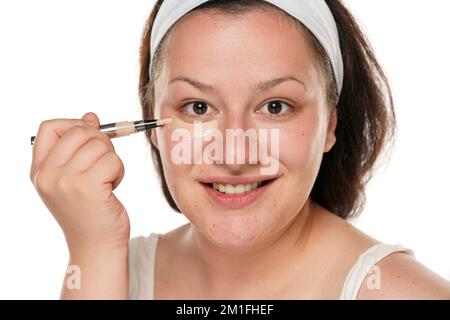 Half portrait of a young smiling chubby woman applyes concealer under ...