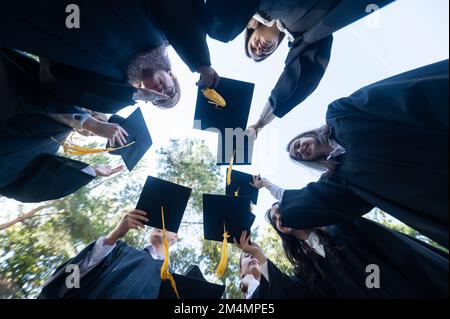 Classmates in graduation gowns toss their hats outdoors. Bottom view ...