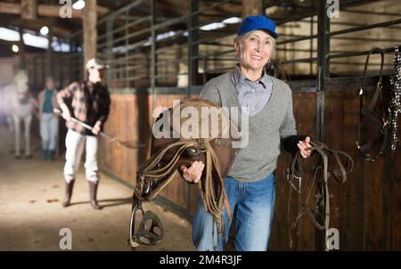 Older female stable keeper carrying horse saddle and harness Stock ...