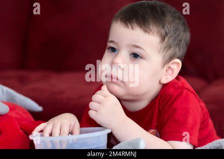adorable boy is watching tv and eating blueberries in the livingroom ...