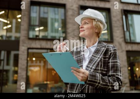 Senior female engineer in suit with white helmet making notes in tablet ...