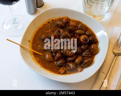 Stewed snails in red sauce served on a white ceramic plate close to the ...