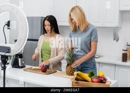 Interracial bloggers cutting fresh fruits near blender and smartphone ...