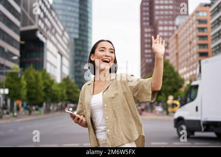 Happy asian girl passing by friend and waving at them on street, saying ...