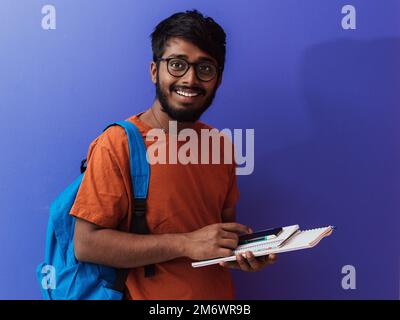Indian student with blue backpack, glasses and notebook posing on gray ...