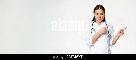 Angry female doctor, asian physician in medical robe and stethoscope, pointing fingers right and ...