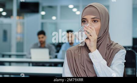 Thirsty millennial indian arabic female worker in hijab holding glass drinking pure mineral ...