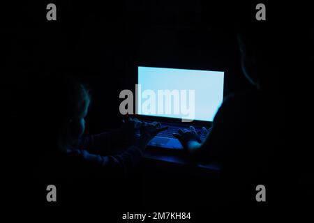 Kids tap on laptop while watch on colored screen in light off room ...