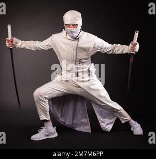A man in the form of a combat ninja poses with two swords on a gray ...