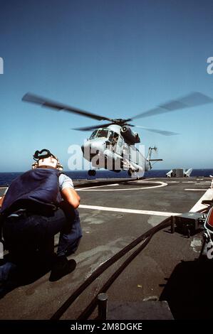 A sailor stands by with wheel chocks and tie-down chains as an SH-2F ...