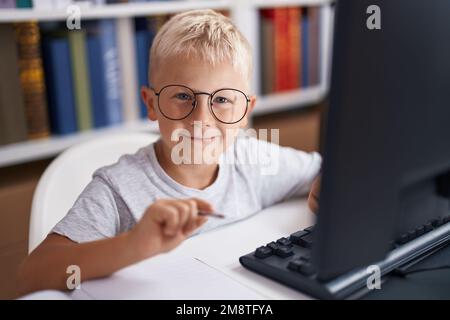 Adorable toddler student using computer sitting on table at classroom ...