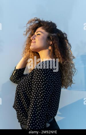 Calm young female in polka dot shirt touching brown curly hair while ...