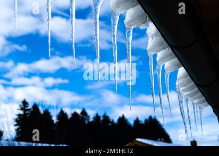 A closeup of ice dams hanging from a roof with cloudy sky blurred ...