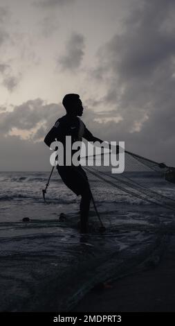 A grayscale shot of a man fishing from the rocky cliff under a cloudy ...