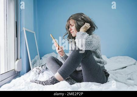 Curly woman with gray sweater sitting on brown sofa and blue blankets ...