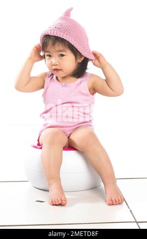 Girl wearing pink vest and pink wool hat, red slippers sitting on white potty holding nappy, 20 ...